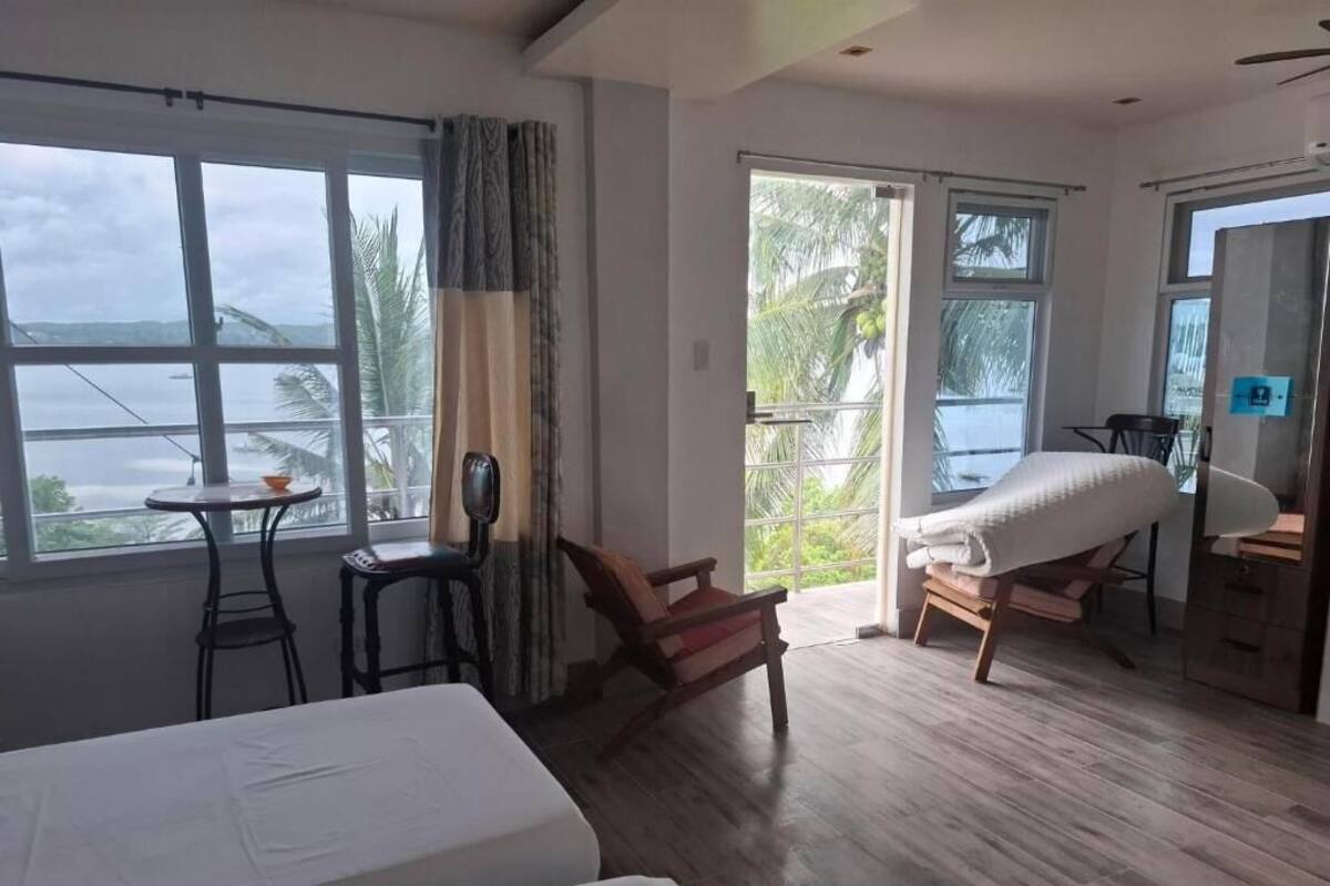 Quarto com cama, uma cadeira, uma mesinha e uma escrivaninha. Há janelas amplas com vista para palmeiras e para o mar.