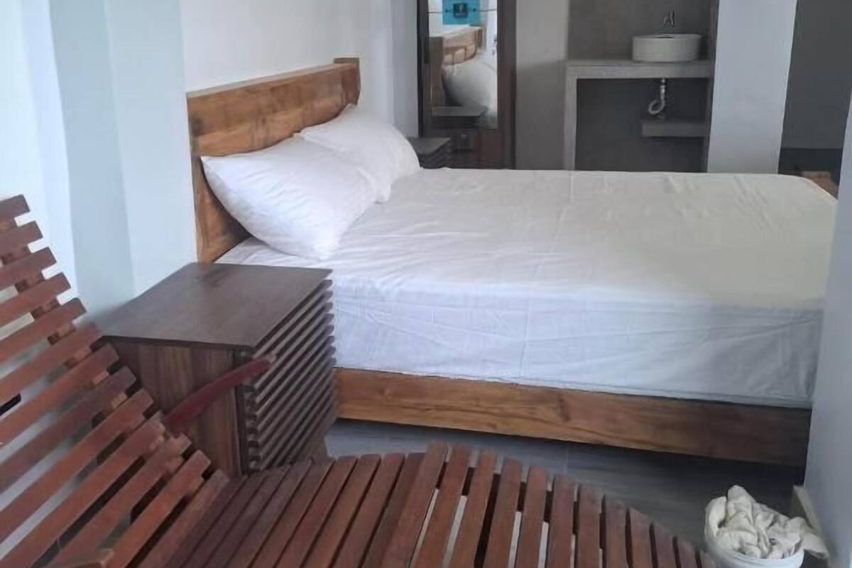 Quarto com cama de madeira, mesa de cabeceira, espelho e um banheiro pequeno com pia.