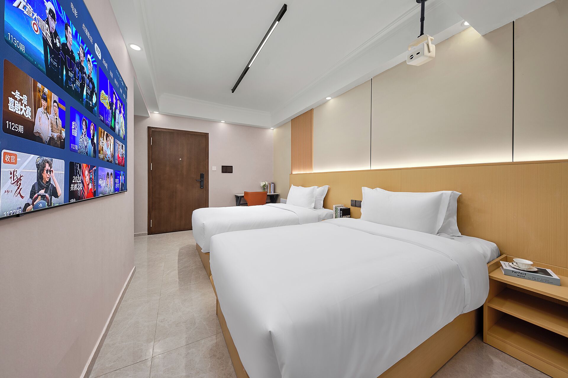 Quarto de hotel com duas camas, uma mesa de cabeceira, escrivaninha e televisão na parede exibindo diversos programas.