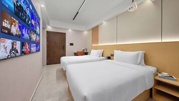 Quarto de hotel com duas camas, uma mesa de cabeceira, escrivaninha e televisão na parede exibindo diversos programas.