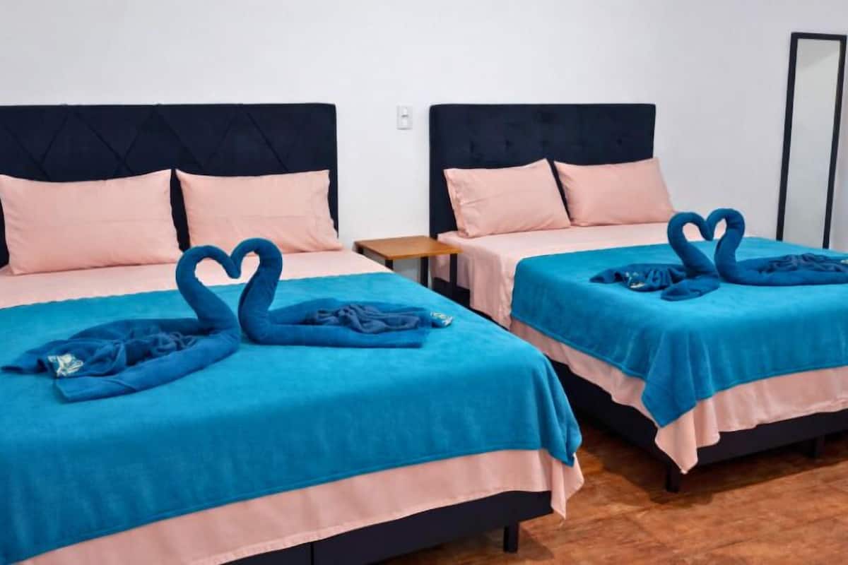 Duas camas de casal com roupa de cama azul e pêssego combinando, dispostas em um quarto com piso de madeira e uma pequena mesa de cabeceira de madeira entre elas.