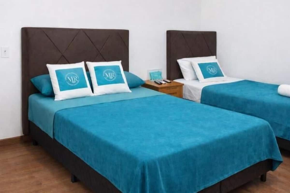 Quarto de hotel com duas camas, cada uma com colcha azul e almofadas combinando, uma mesinha de cabeceira de madeira e uma parede branca.