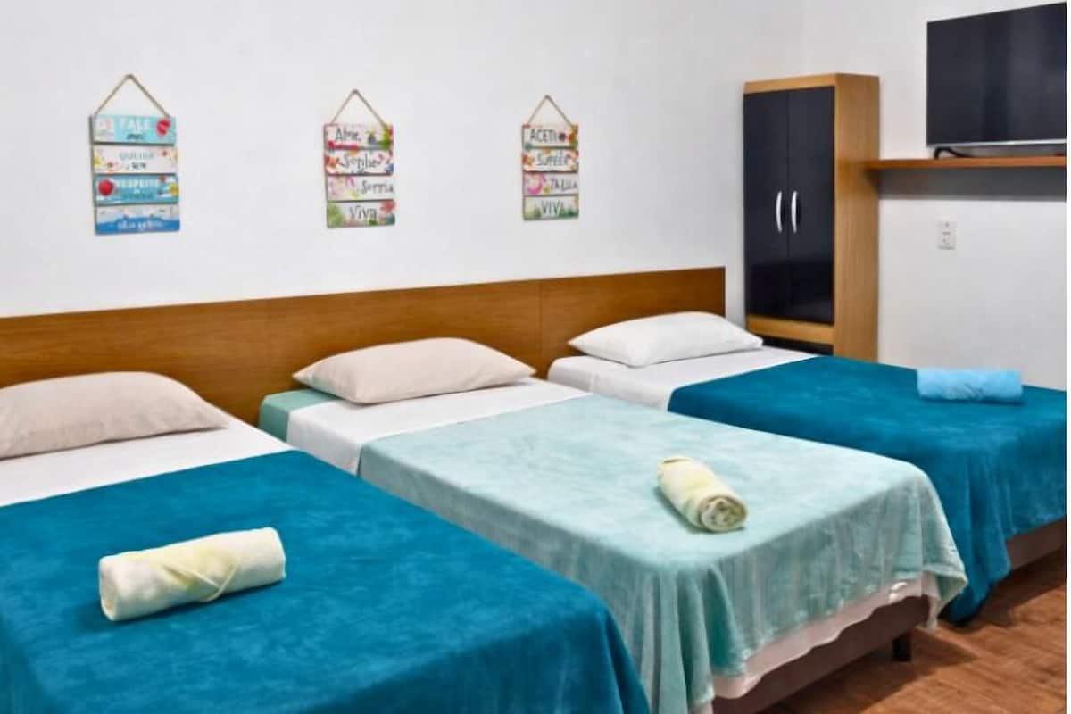Quarto de hotel com três camas de solteiro, cabeceira de madeira, guarda-roupa e detalhes decorativos nas paredes.