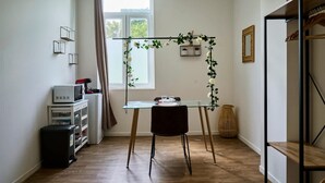 Quarto compacto com mesa de vidro, uma cadeira preta, pequeno armário, micro-ondas, janela com cortinas e piso de madeira.