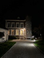 Um edifício grande e histórico com uma torre imponente, uma galeria coberta e uma entrada com acesso por alamedas.