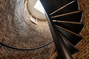 Escadaria em espiral com claraboia, paredes de tijolo e plataforma de madeira no topo.
