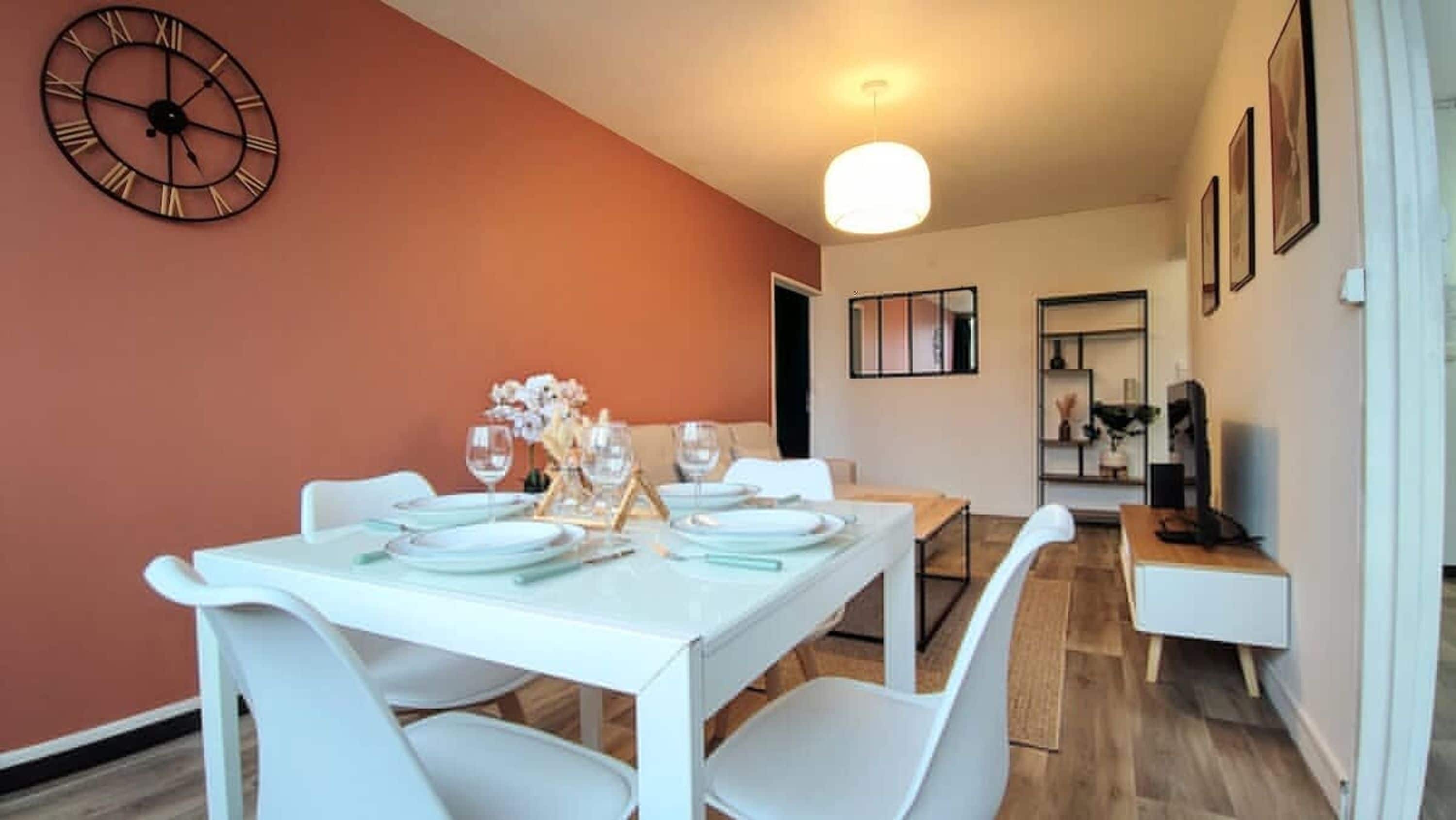 Uma sala de jantar com uma mesa branca posta para quatro pessoas, um relógio na parede e um corredor que dá para uma cozinha.