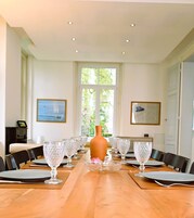 Uma sala de jantar com uma longa mesa de madeira posta para uma refeição, ladeada por cadeiras, com um vaso e taças de cristal sobre a mesa.