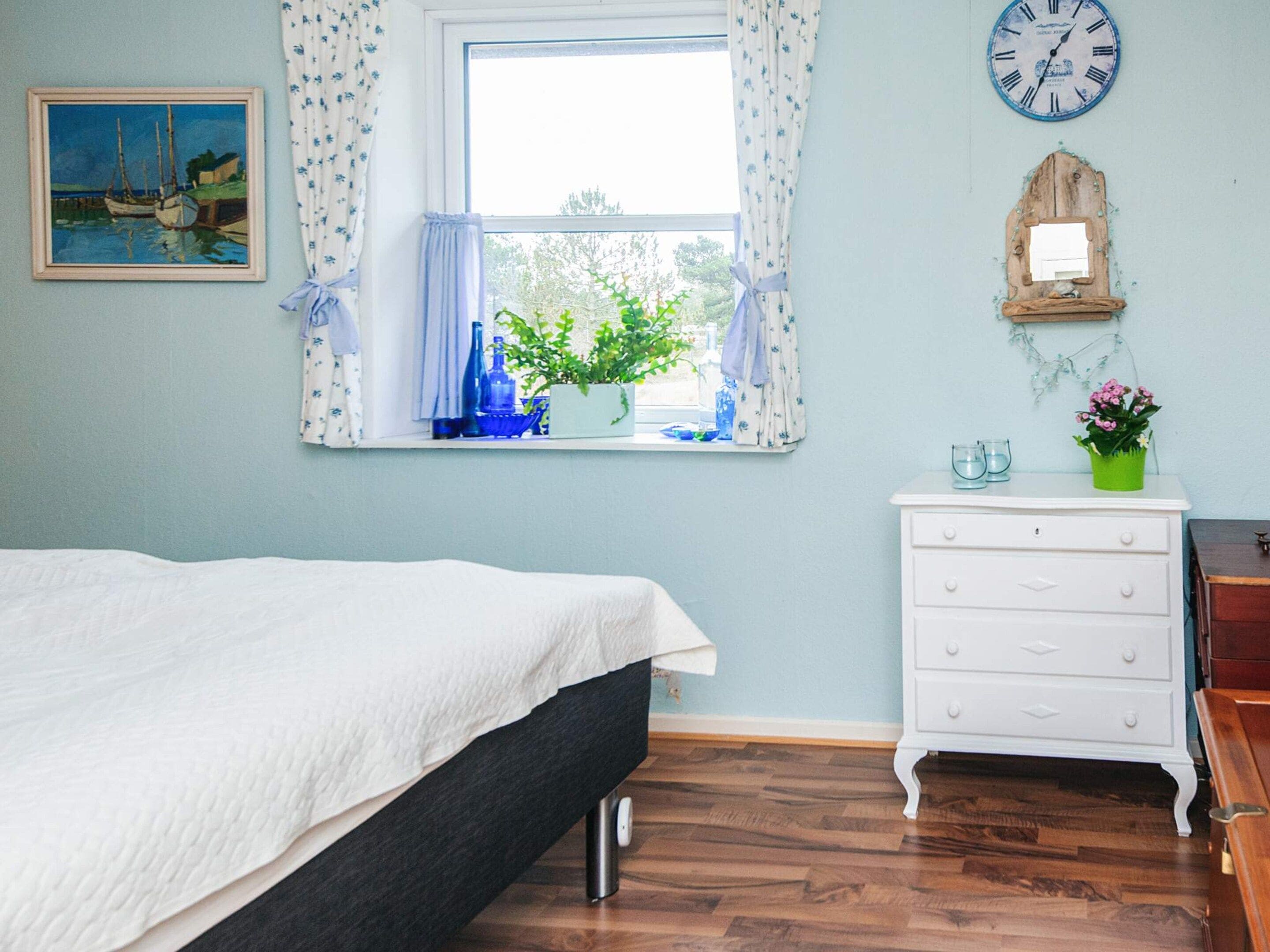 Quarto com cama, penteadeira, janela com cortinas, relógio, quadro e planta em vaso.