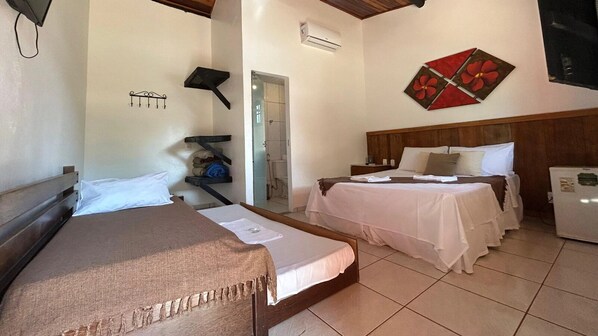 Quarto de hotel com duas camas, cabeceira de madeira, uma mesa pequena, ar condicionado fixado na parede e porta para um banheiro.