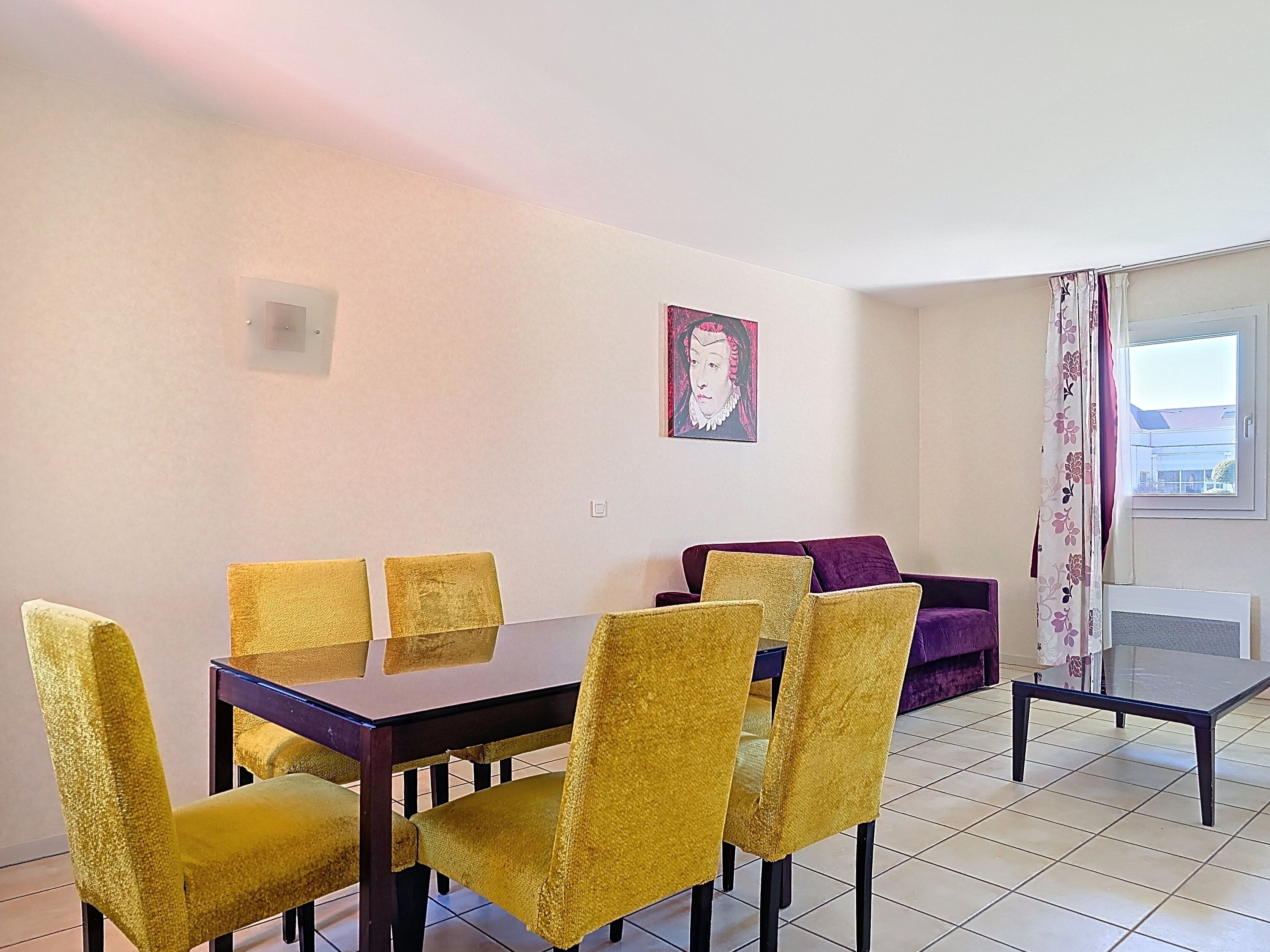Uma sala de jantar com mesa e cadeiras, um sofá roxo e uma janela com cortinas.