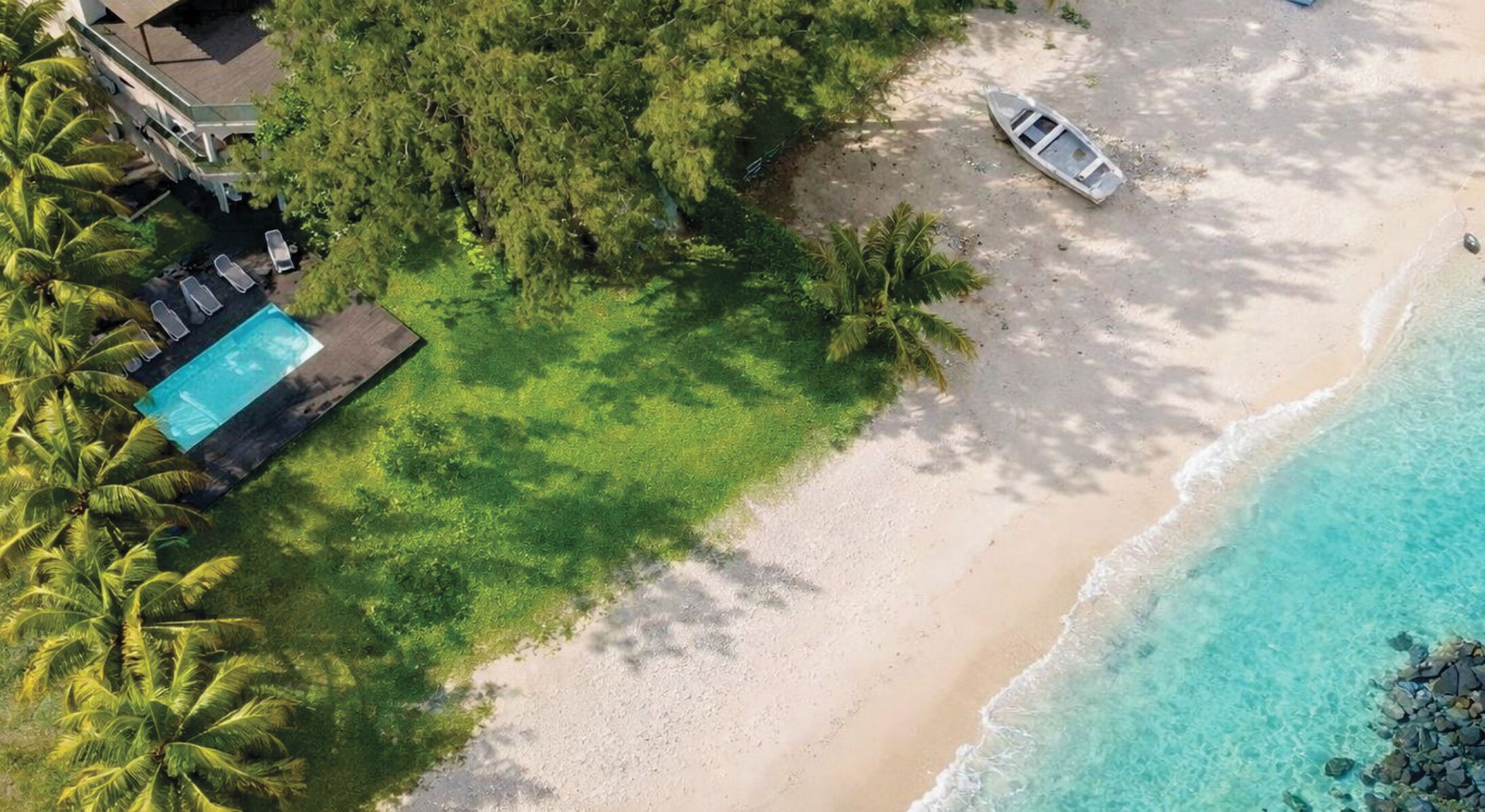 Vista aérea de uma praia com piscina, barco e vegetação exuberante.