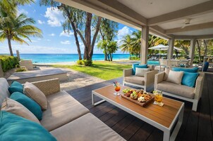 Uma área de estar coberta ao ar livre com vista para a praia e o oceano, equipada com sofás e cadeiras, e uma mesa com petiscos e bebidas.