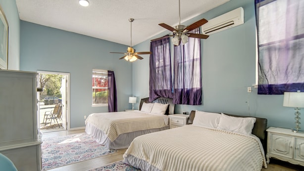 Quarto com duas camas, ventilador de teto, ar condicionado, janela com cortinas roxas e vista para o exterior.