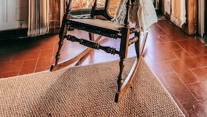 Uma cadeira de balanço de madeira em um quarto com uma janela grande, uma cadeira trançada e um tapete no chão.