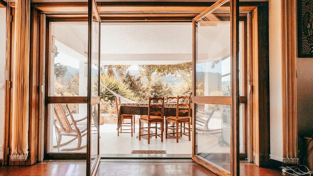Quarto com porta de vidro deslizante que dá para uma área de estar ao ar livre, com cadeiras e mesa de madeira.