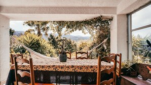 Uma área de refeições com mesa e cadeiras, uma rede e vista para montanhas e árvores.