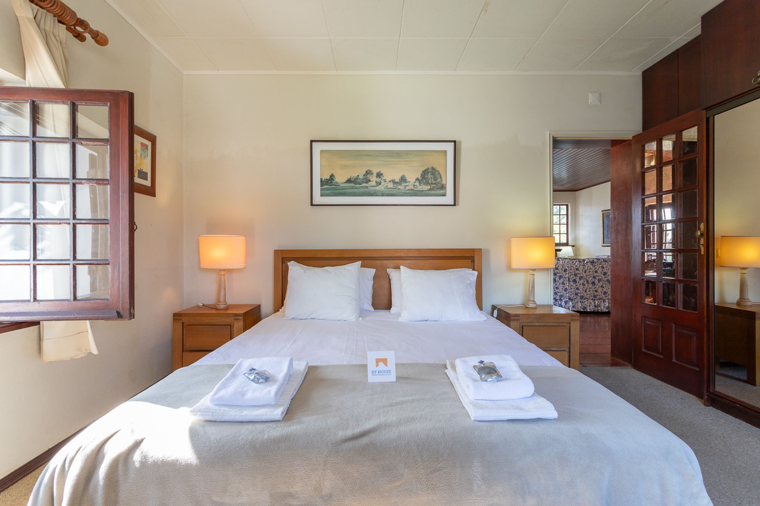 Quarto de hotel com uma cama grande, duas mesas de cabeceira com luminárias, um quadro na parede e um espelho na porta.