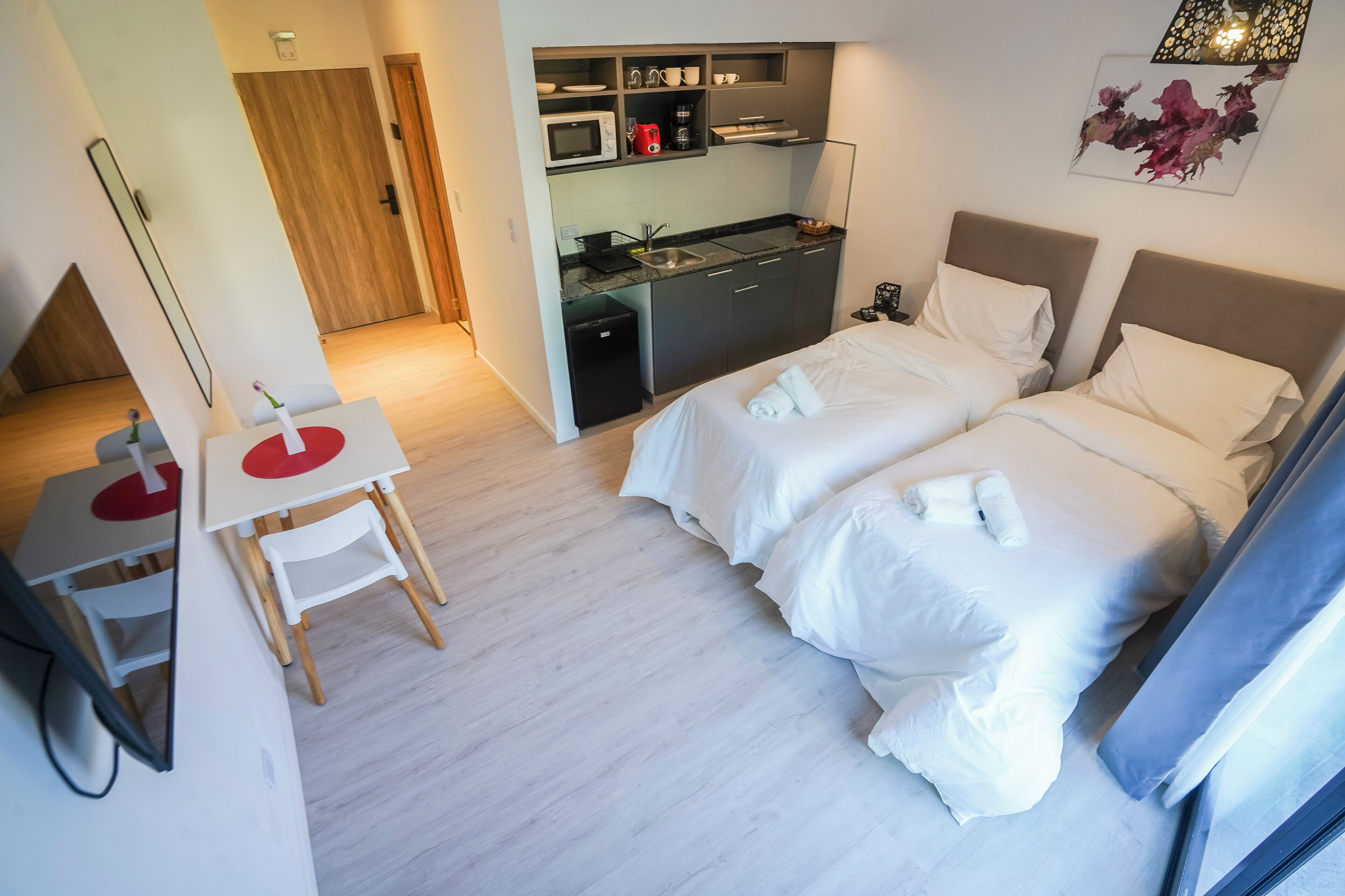 Quarto de hotel com duas camas, cozinha compacta, uma pequena mesa com cadeiras e um espelho.