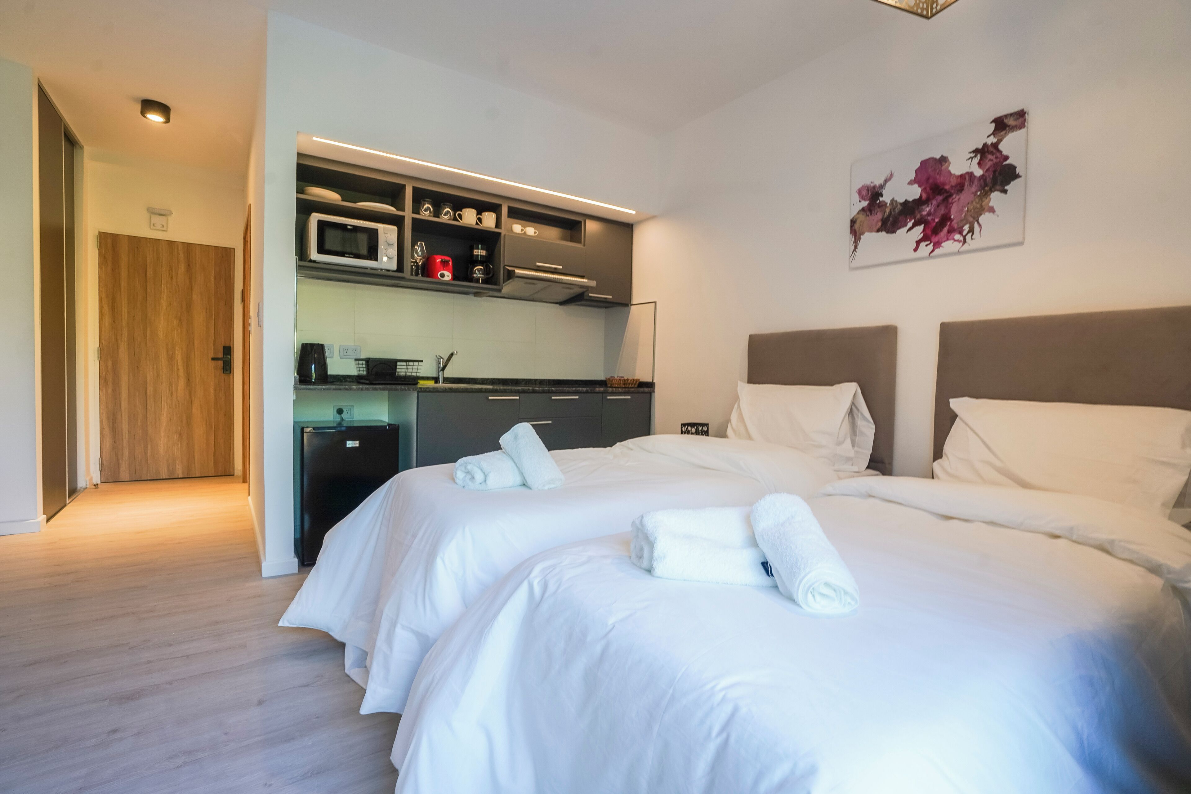 Quarto de hotel com duas camas, uma cozinha compacta e uma porta de madeira.