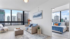 Uma sala de estar moderna com sofá, banquinho e mesa de centro, uma cama com cobertor azul e vista para a cidade através de grandes janelas.