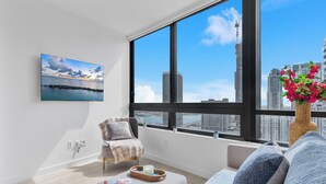 Uma sala de estar moderna com uma janela grande, um sofá, uma poltrona, uma mesinha com livros e velas, e um quadro de uma paisagem costeira.