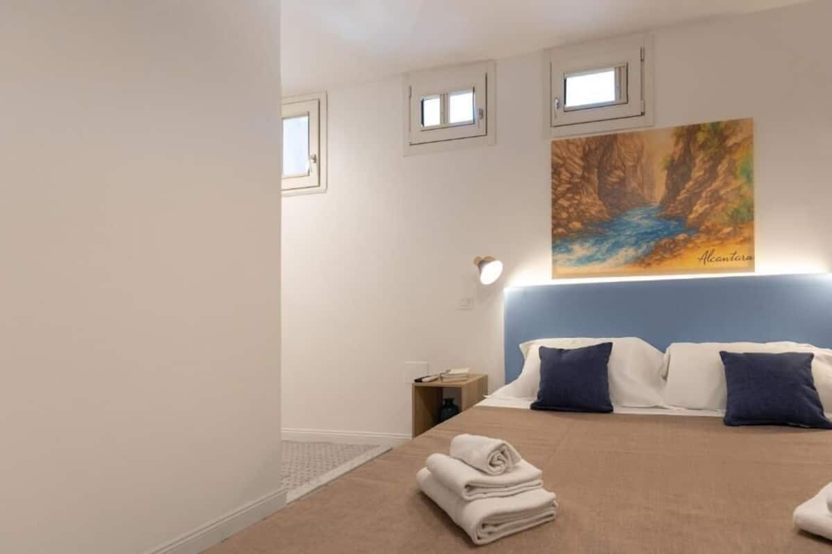 Quarto de hotel com cama, mesa de cabeceira, um quadro e cabeceira.