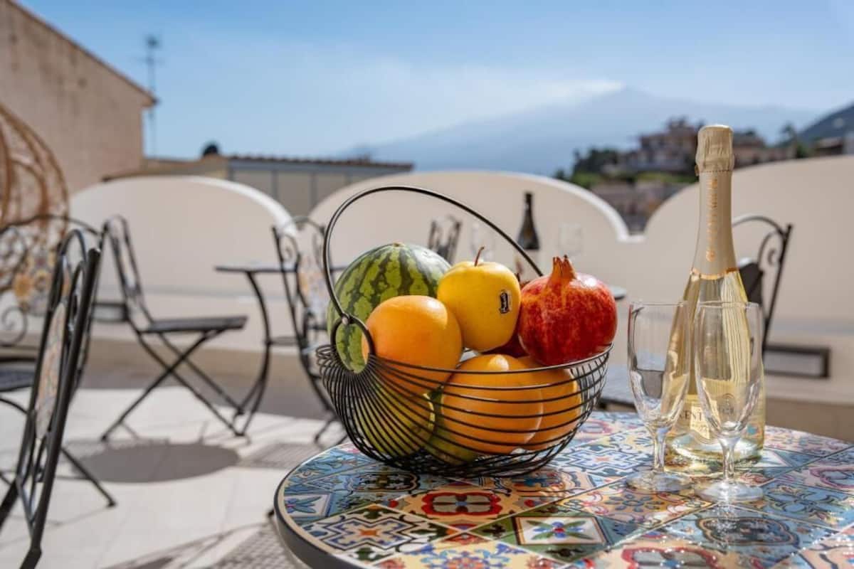 Uma mesa com uma cesta de frutas, uma garrafa de champanhe e copos sobre uma toalha de mesa estampada.