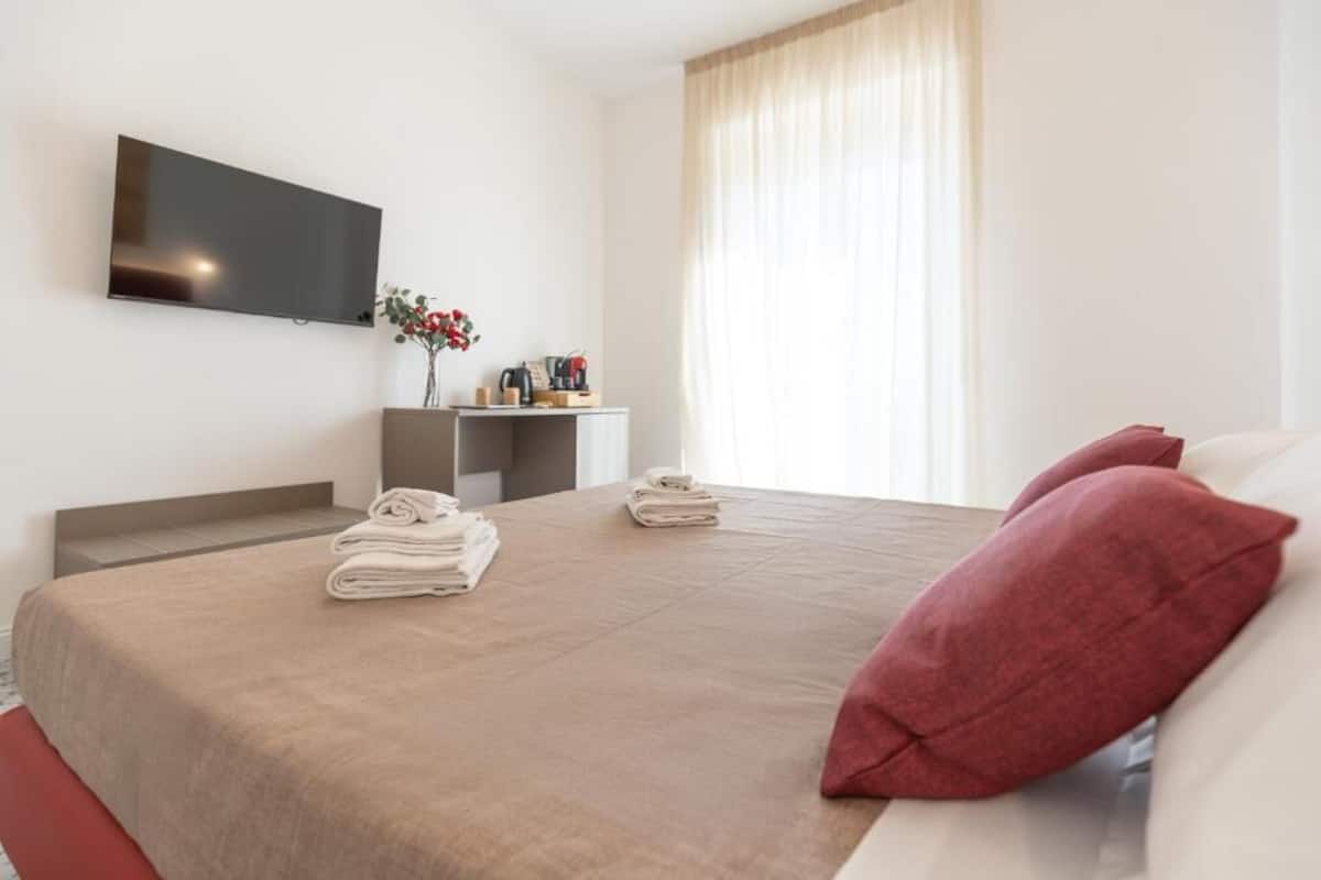 Quarto de hotel com cama, televisão na parede, mesa de cabeceira com um vaso de flores e janela com cortinas.