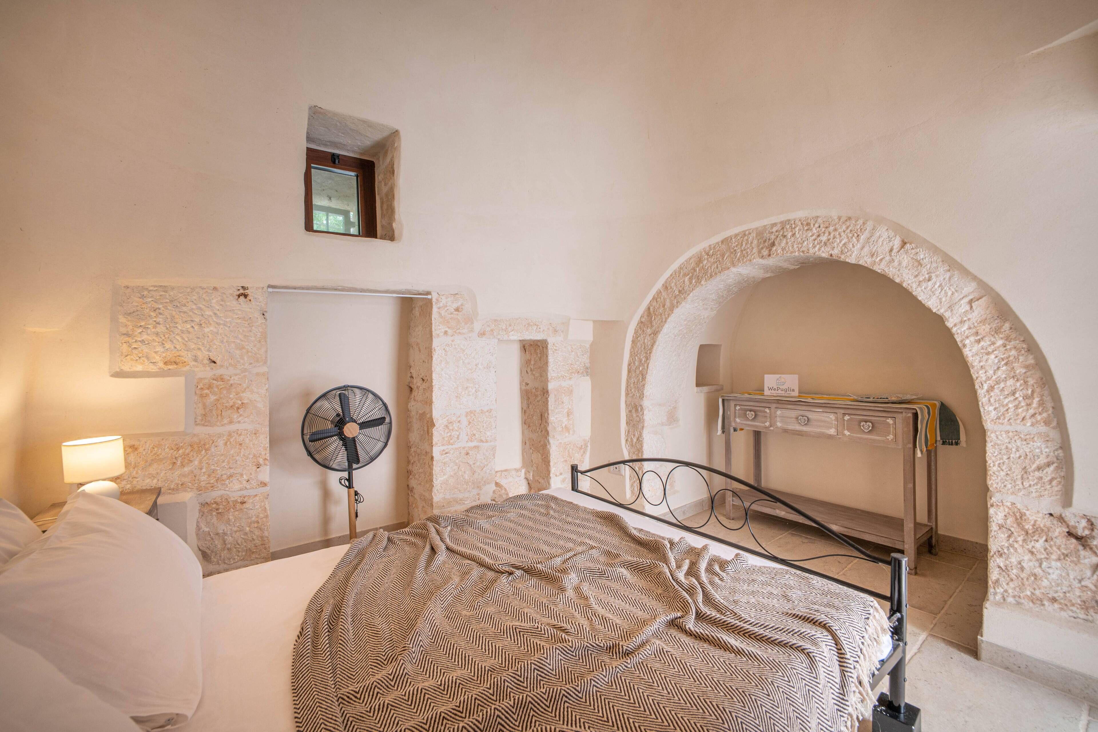 Quarto com cama, mesa de cabeceira, abajur, ventilador e parede de pedra em arco.