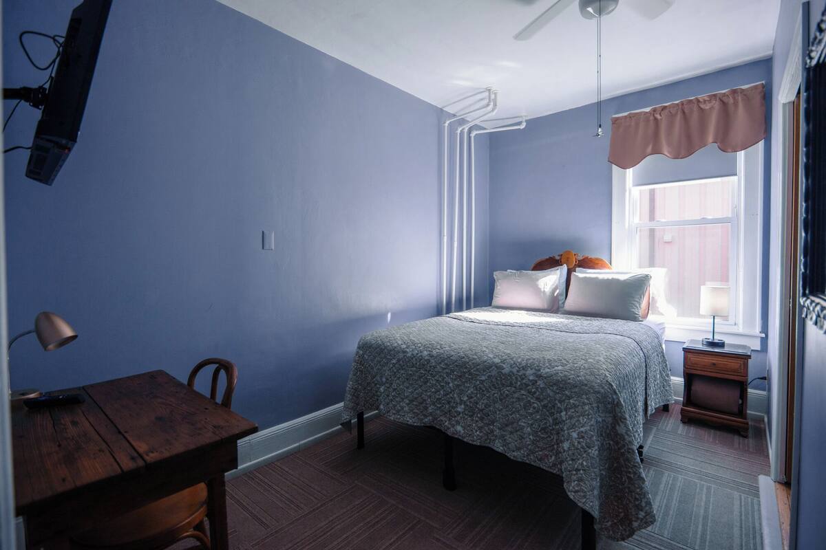 Quarto com cama, escrivaninha, cadeira, janela com cortinas e ventilador de teto.