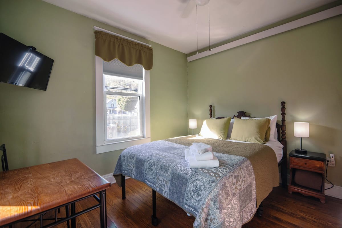 Quarto com cama, mesas de cabeceira, escrivaninha, janela com cortinas e televisão na parede.
