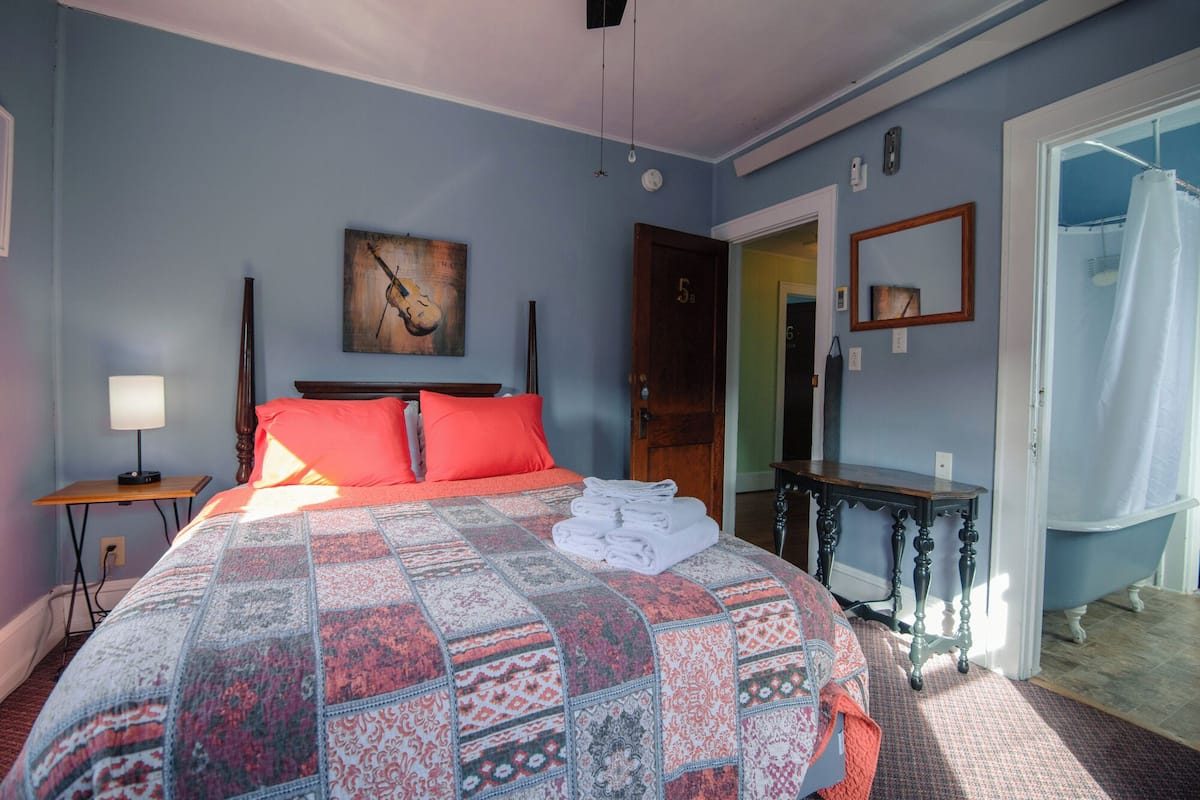 Quarto com cama, mesa de cabeceira, relógio na parede, porta e banheiro visível através de uma porta aberta.
