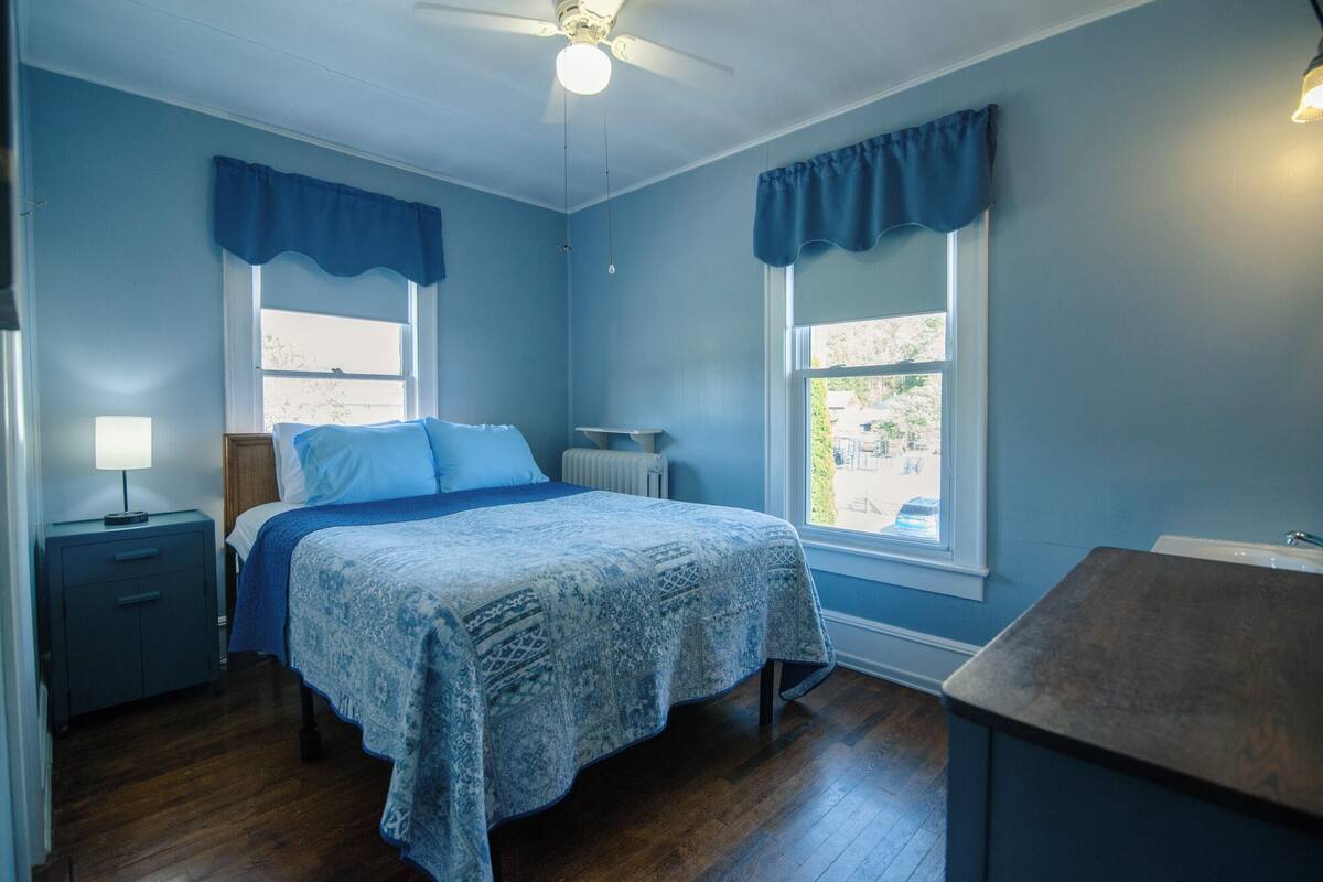Quarto com cama, mesa de cabeceira, abajur, janela e ventoinha de teto.