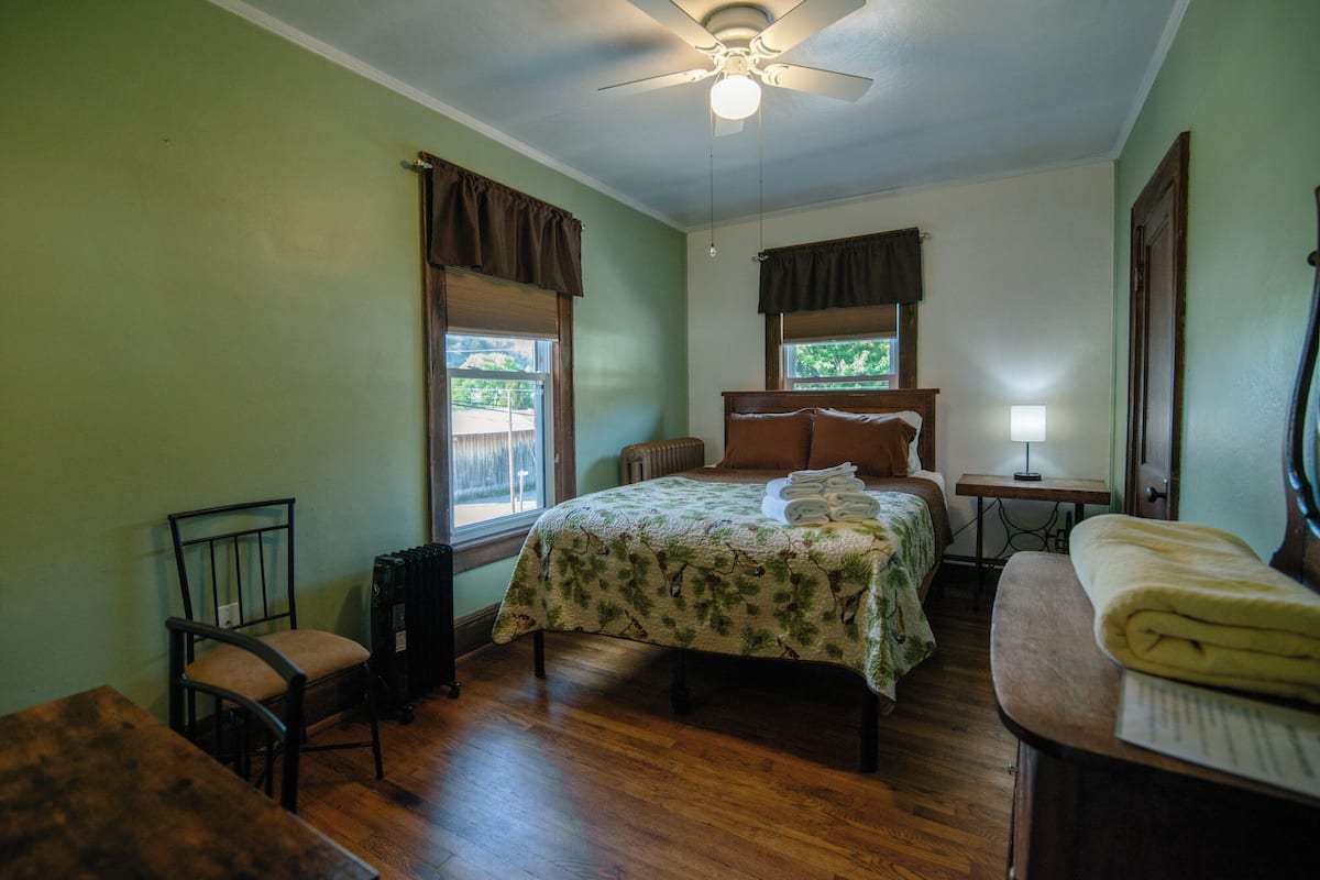 Quarto com cama, cadeira, mesa, ventilador de teto e janela.