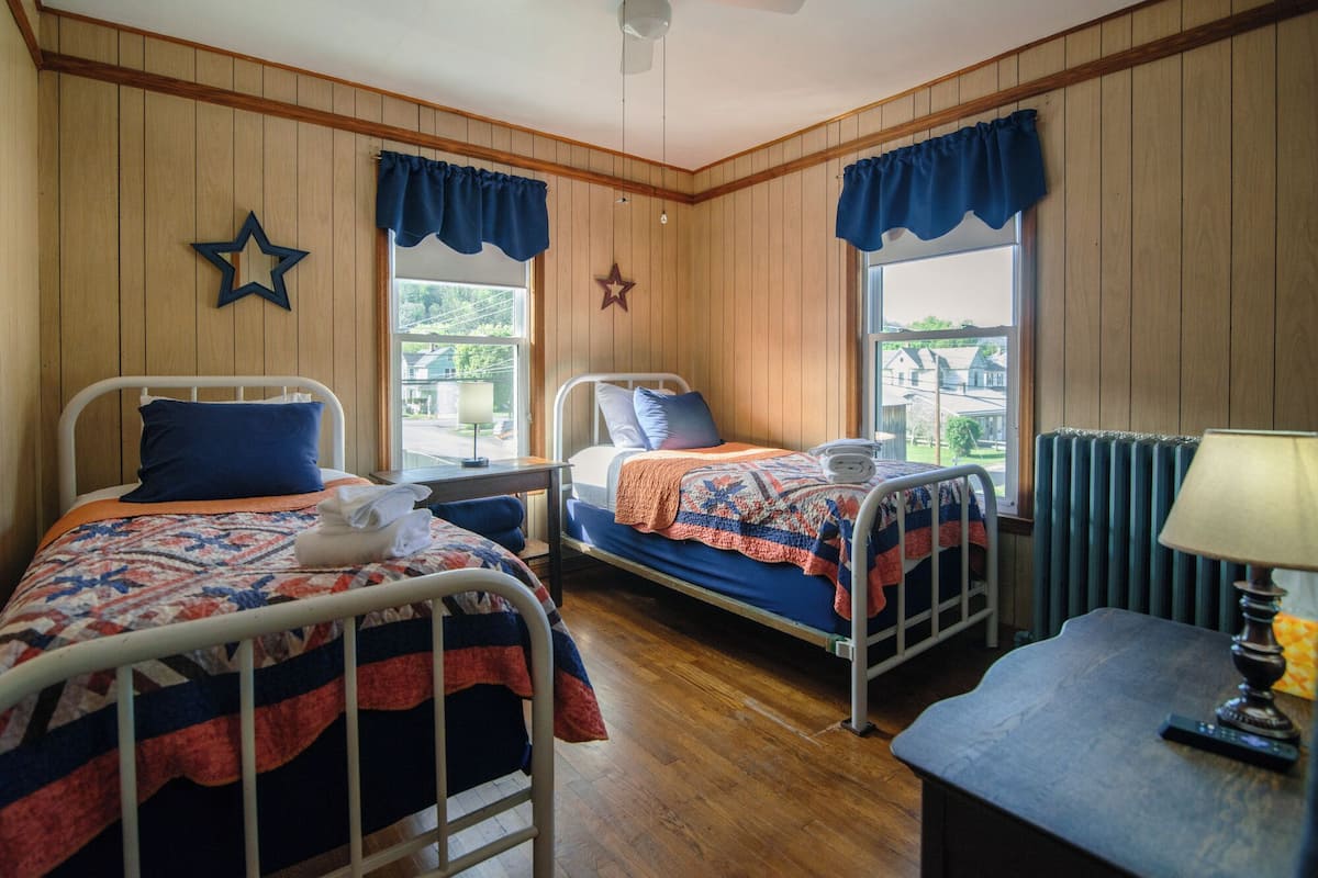 Quarto com duas camas, paredes de madeira, detalhes de estrelas e vista para casas pelas janelas.