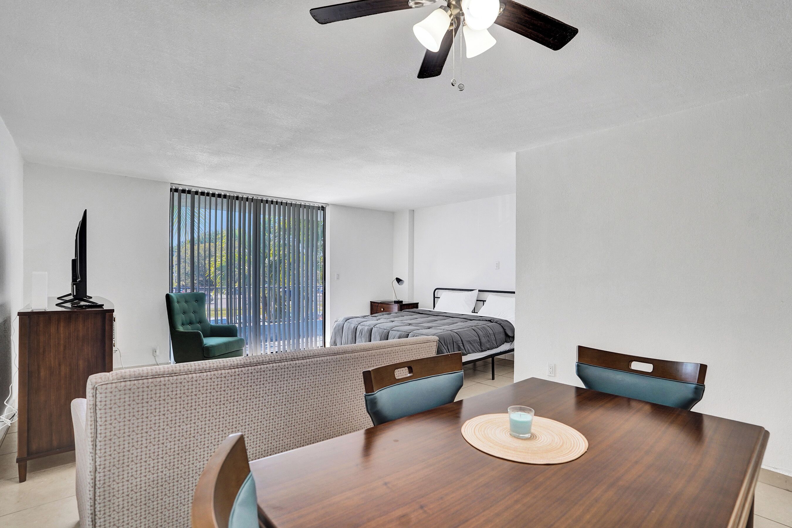 Quarto de hotel moderno com uma mesa de jantar, cadeiras, cama, televisão e janela com persianas.