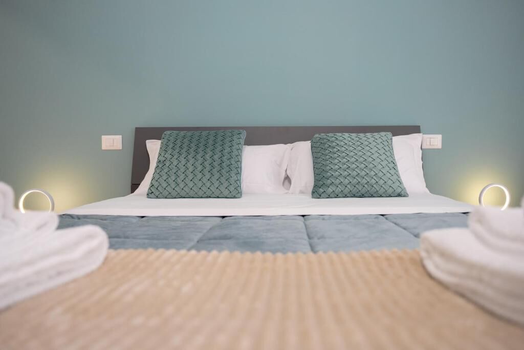 Cama bem arrumada com travesseiro branco e verde estampado, cabeceira cinza e parede azul clara.