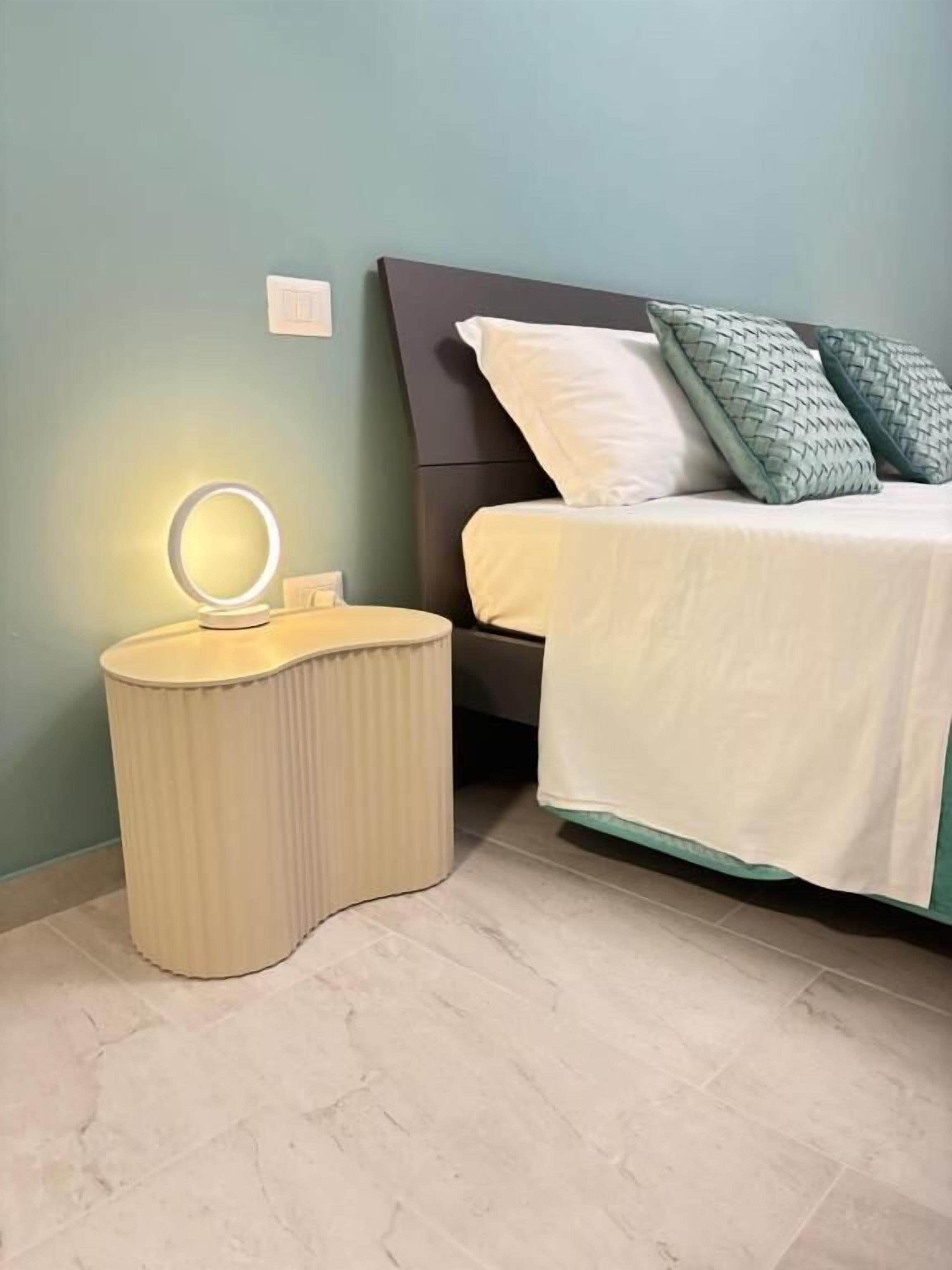 Um quarto com uma cama, uma mesa de cabeceira com um espelho redondo e uma tomada na parede.
