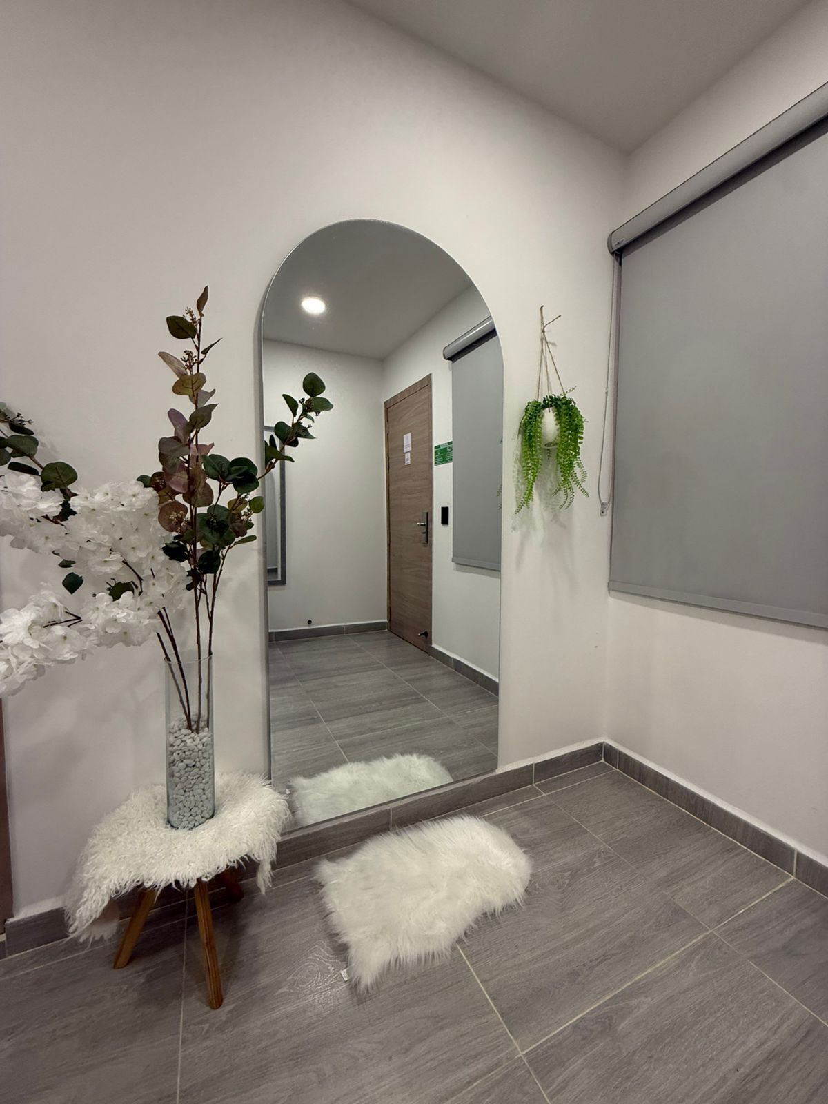 Um corredor com um tapete branco e fofo, um vaso alto com flores brancas e uma planta pendente verde.