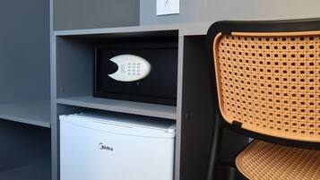 Uma geladeira compacta com painel de controle, uma cadeira de vime e uma parede cinza com tomada.