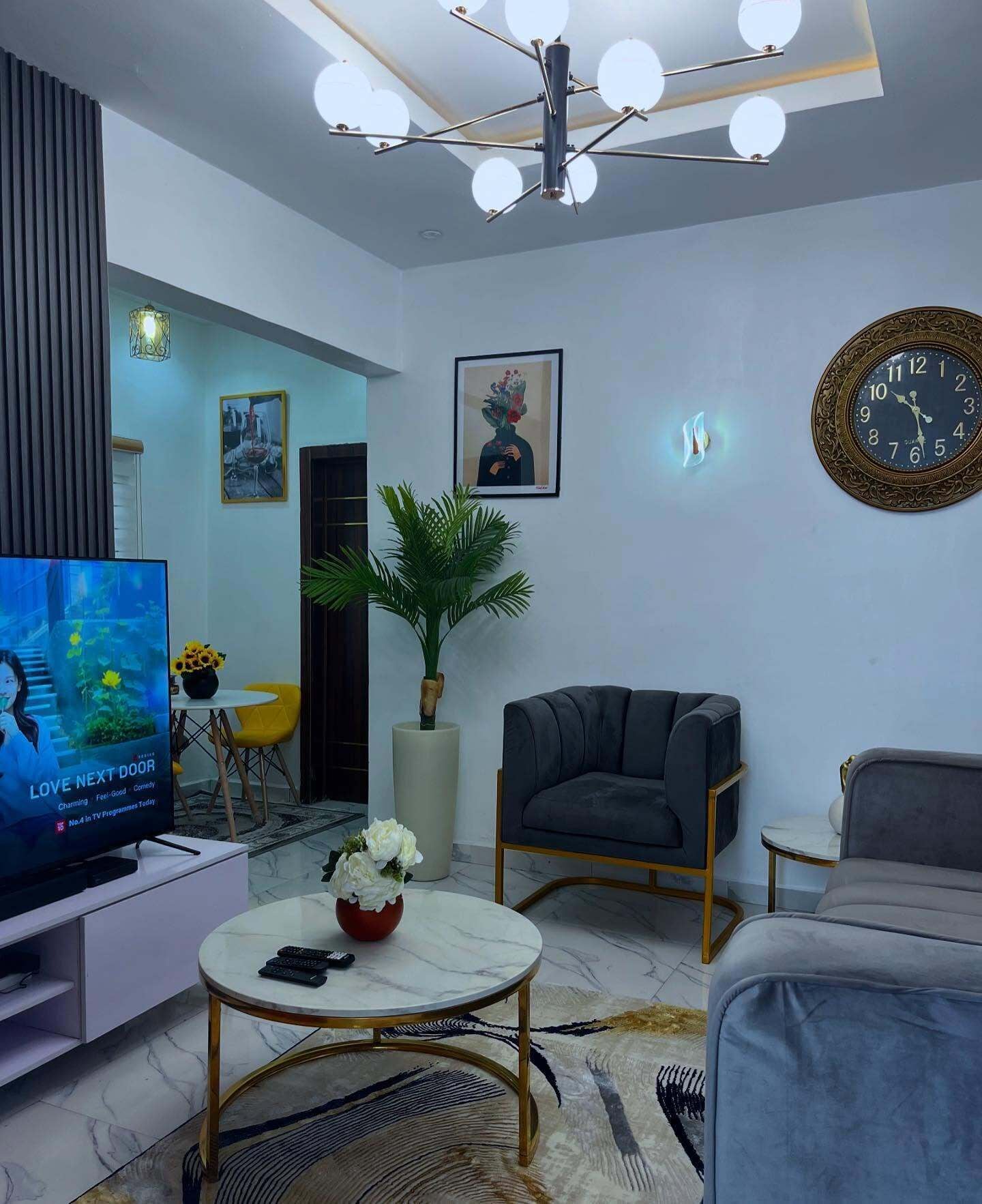 Uma sala de estar moderna com televisão de tela plana, um sofá cinza, uma mesa redonda com um vaso, uma planta em vaso, um relógio de parede e um lustre.