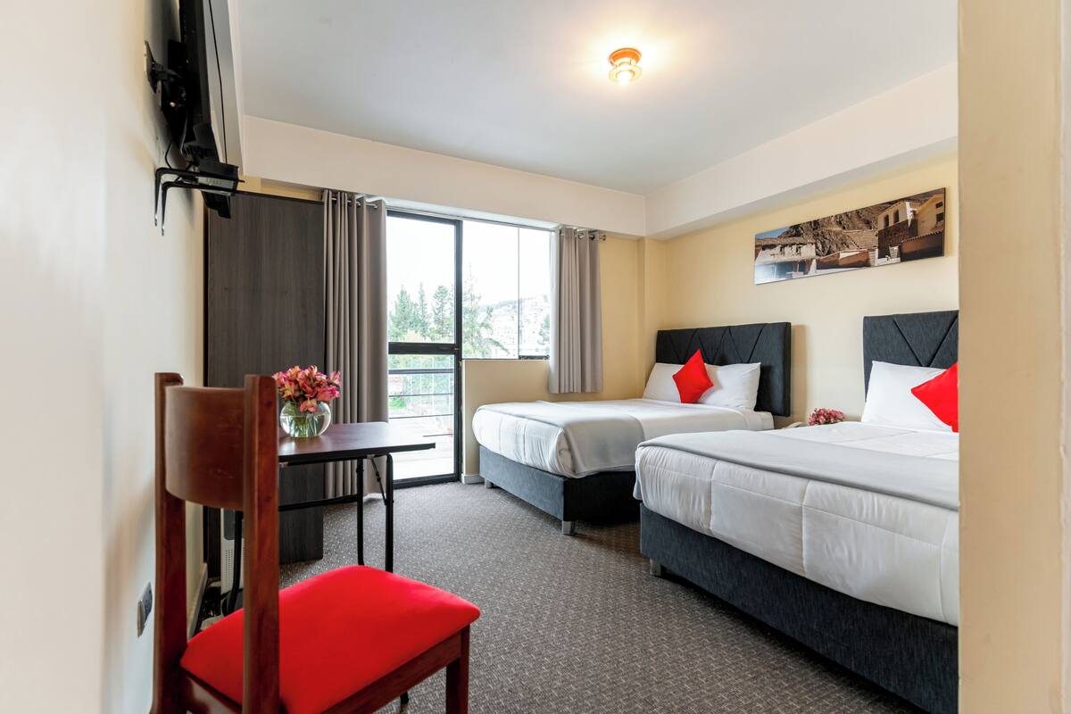 Quarto de hotel com duas camas, uma escrivaninha, uma cadeira e uma janela com cortinas.