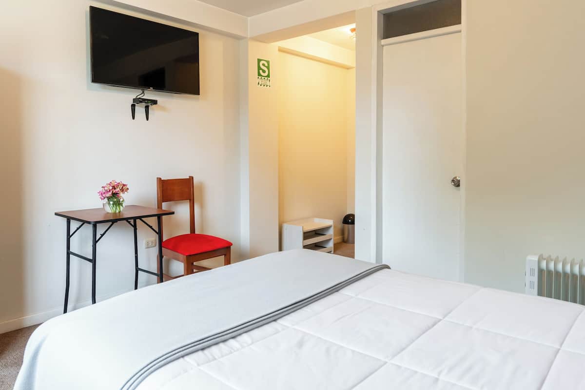 Quarto de hotel com cama, televisão na parede, escrivaninha com cadeira e radiador.