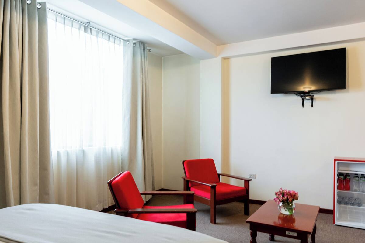 Quarto de hotel com uma cama, duas cadeiras vermelhas, uma mesinha com um vaso, uma TV de tela plana na parede e um escaldem para sapatos.