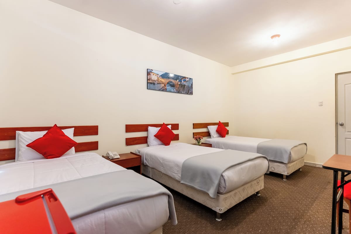 Quarto de hotel com três camas, cada uma com cabeceira de madeira e almofadas vermelhas, uma mesinha e um quadro na parede.