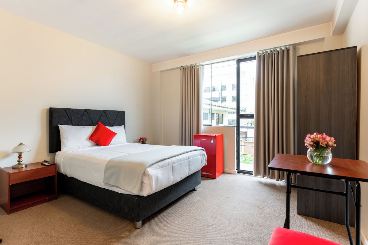 Quarto de hotel com uma cama, uma mesinha de cabeceira, um armário vermelho, uma mesa com um vaso de flores e vista para o exterior por uma grande janela.