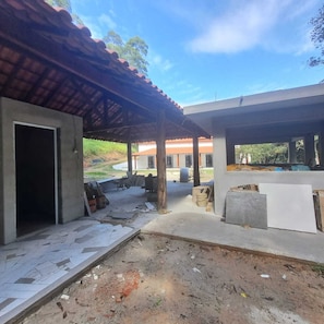 Área externa coberta com piso de concreto, uma porta e um edifício em construção ao fundo.