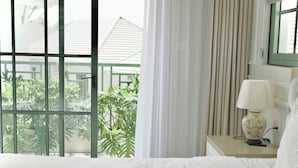 Quarto de hotel com cama, mesa de cabeceira com abajur, janela com cortinas e vista para o verde lá fora.