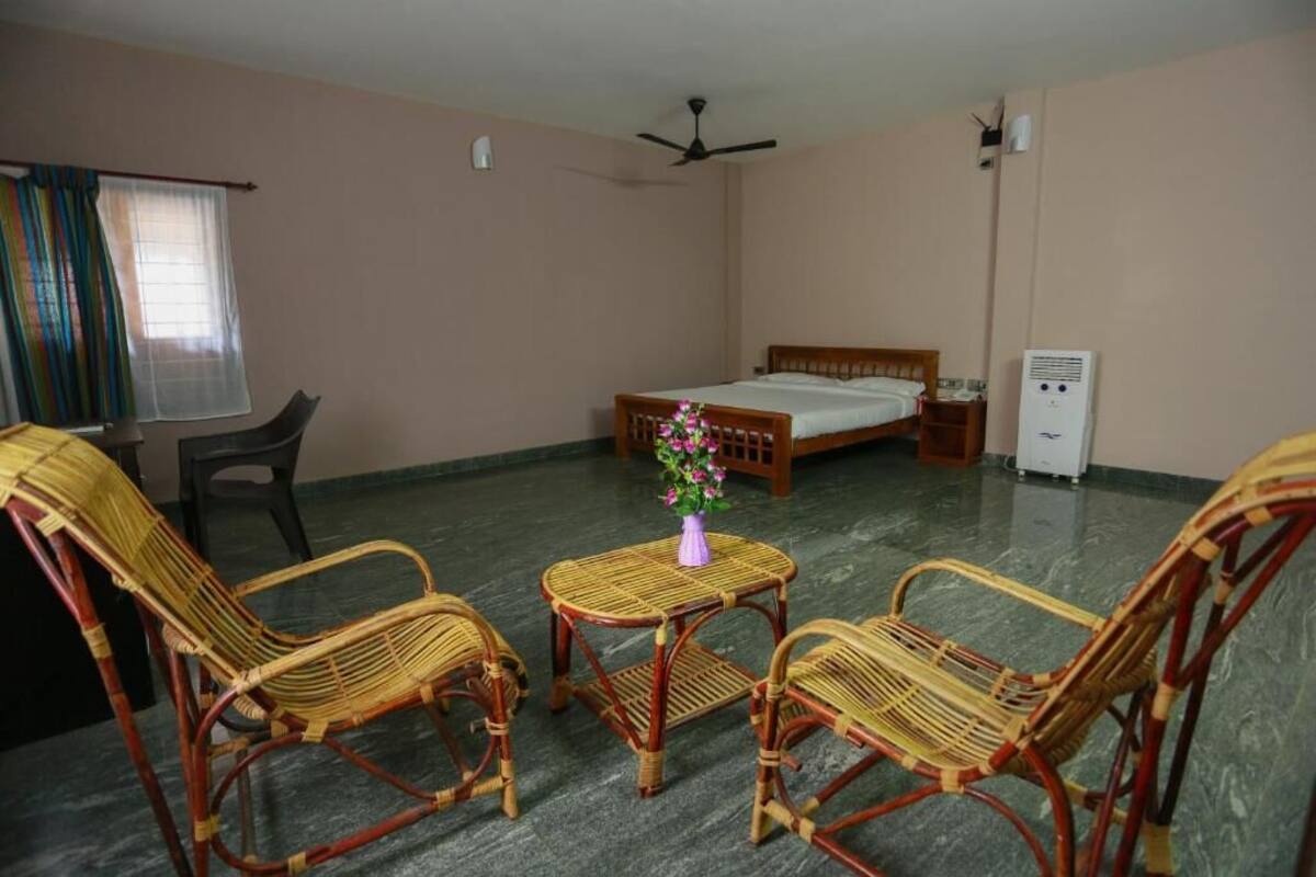 Quarto com uma cama, duas cadeiras de vime, uma mesinha com um vaso, um ventilador de teto e um aquecedor.
